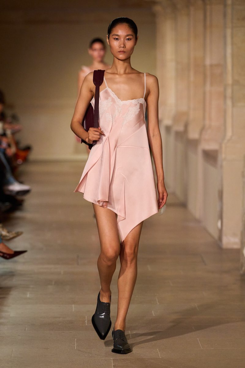 Model wearing a Victoria Beckham spring-summer 2026 blush pink asymmetrical slip dress with lace detailing and black pointed oxford shoes