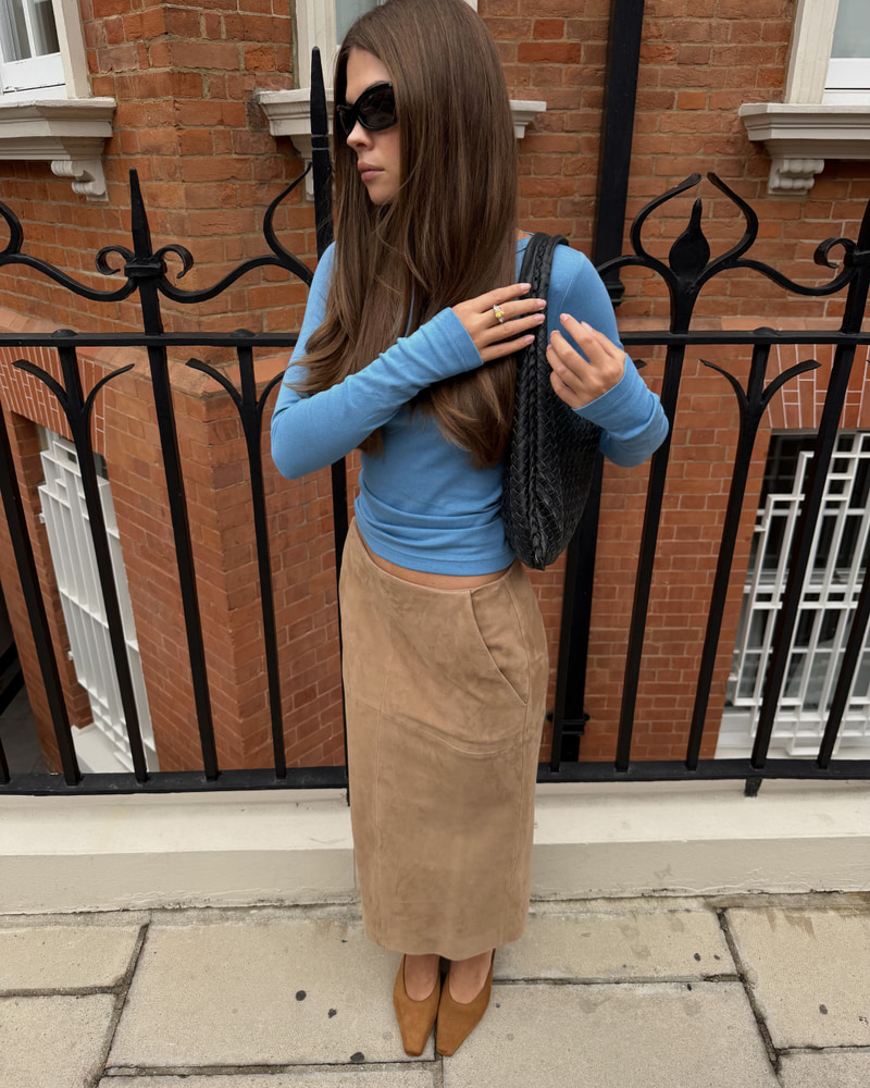 Chic street style featuring a fitted blue long-sleeve top paired with a soft beige suede midi skirt and sultry pointed-toe heels%2C complemented by oversized sunglasses and a textured bag