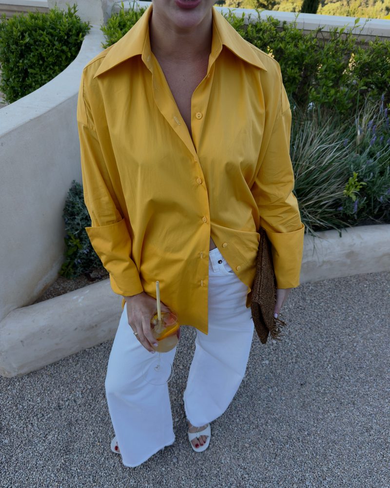 Oversized mustard yellow button-down shirt styled with white wide-leg pants%2C woven clutch%2C and heeled sandals for a chic resort outfit
