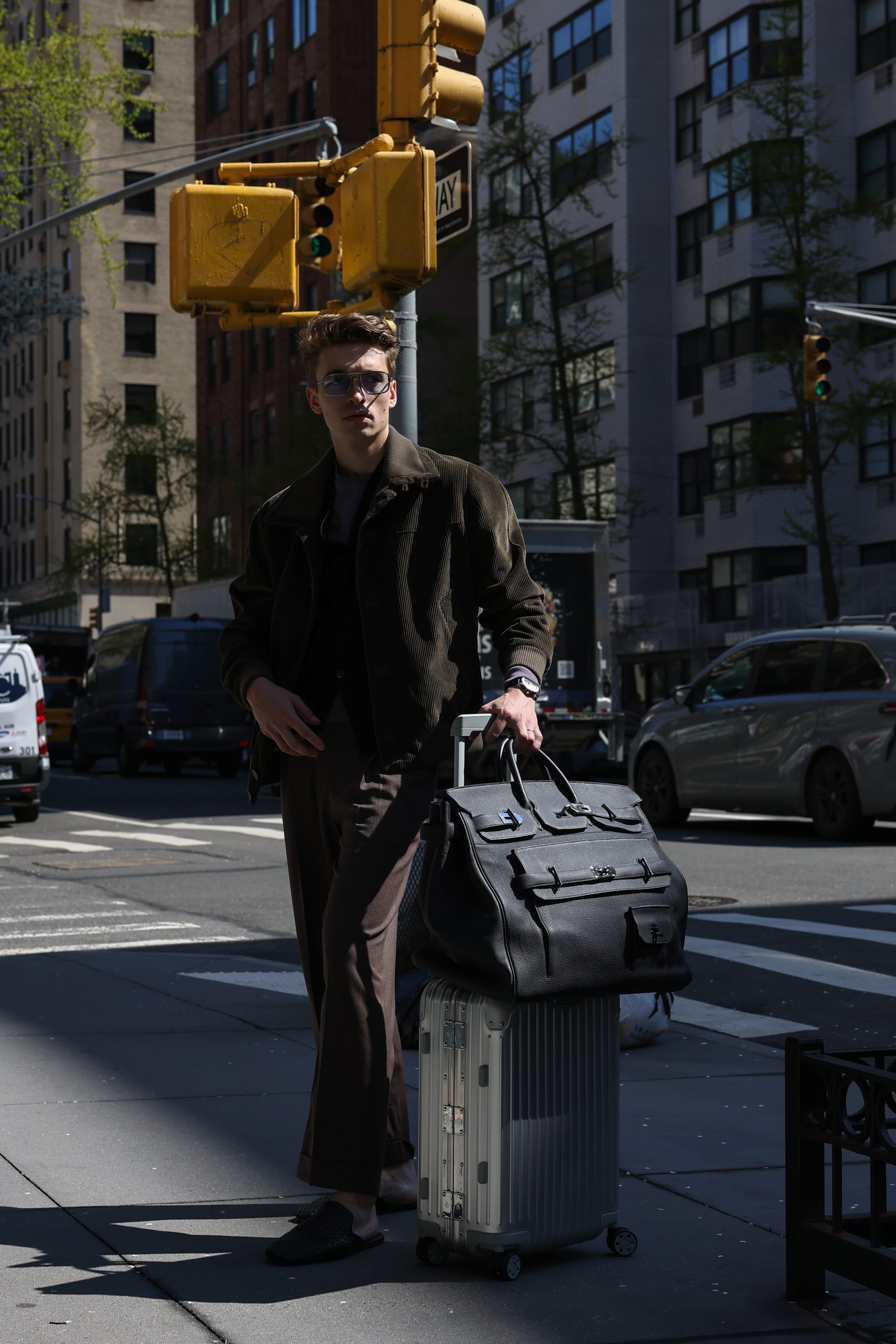 Stylish young man in a dark corduroy jacket and brown trousers stands confidently beside a sleek silver suitcase and a black leather duffel bag%2C set against an urban backdrop