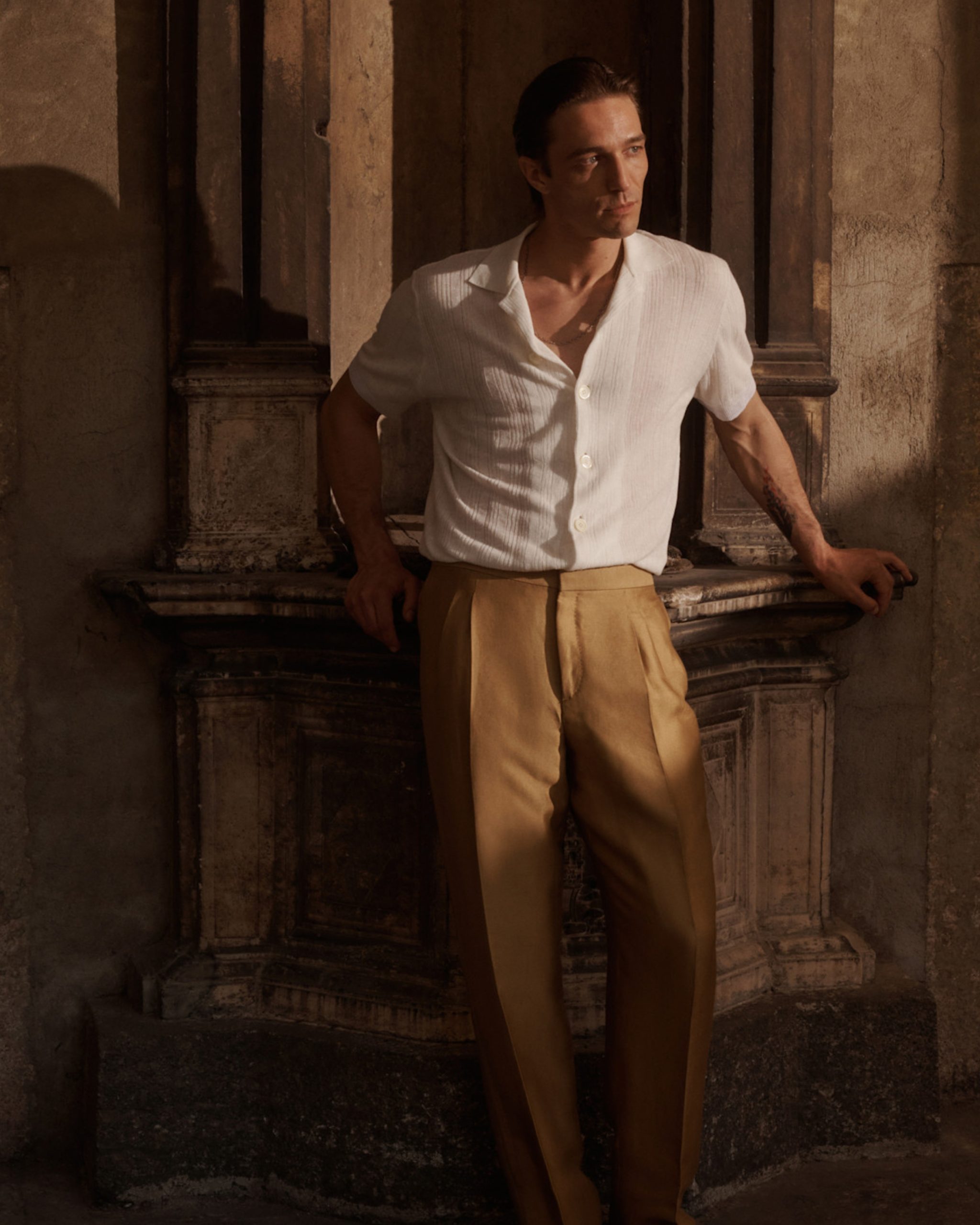 Man wearing Brioni short-sleeve white textured shirt and high-waisted camel trousers in a warm%2C historic setting.