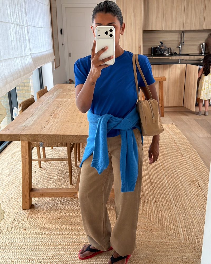 Person taking a selfie in a modern kitchen%2C wearing a blue shirt and brown pants with a blue sweater tied around the waist. A child in a yellow dress is in the background.