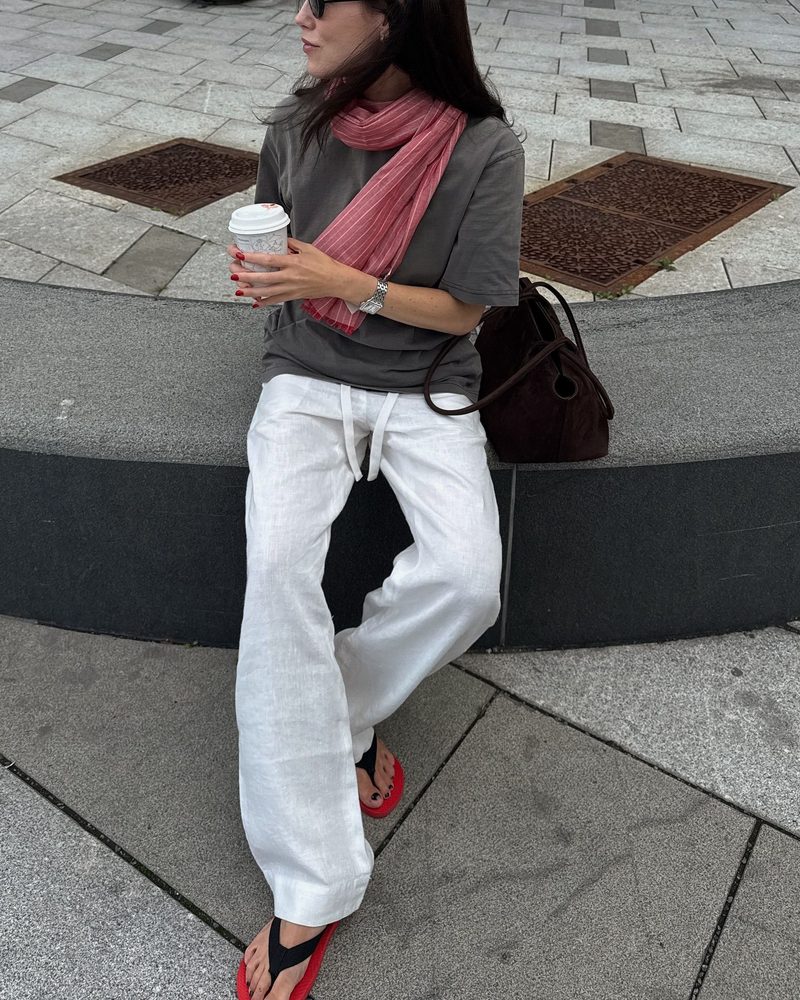 Woman wearing a charcoal gray t-shirt%2C white linen wide-leg pants%2C pink striped scarf%2C black sunglasses%2C and red flip-flops sitting outdoors with a coffee cup and dark brown suede handbag