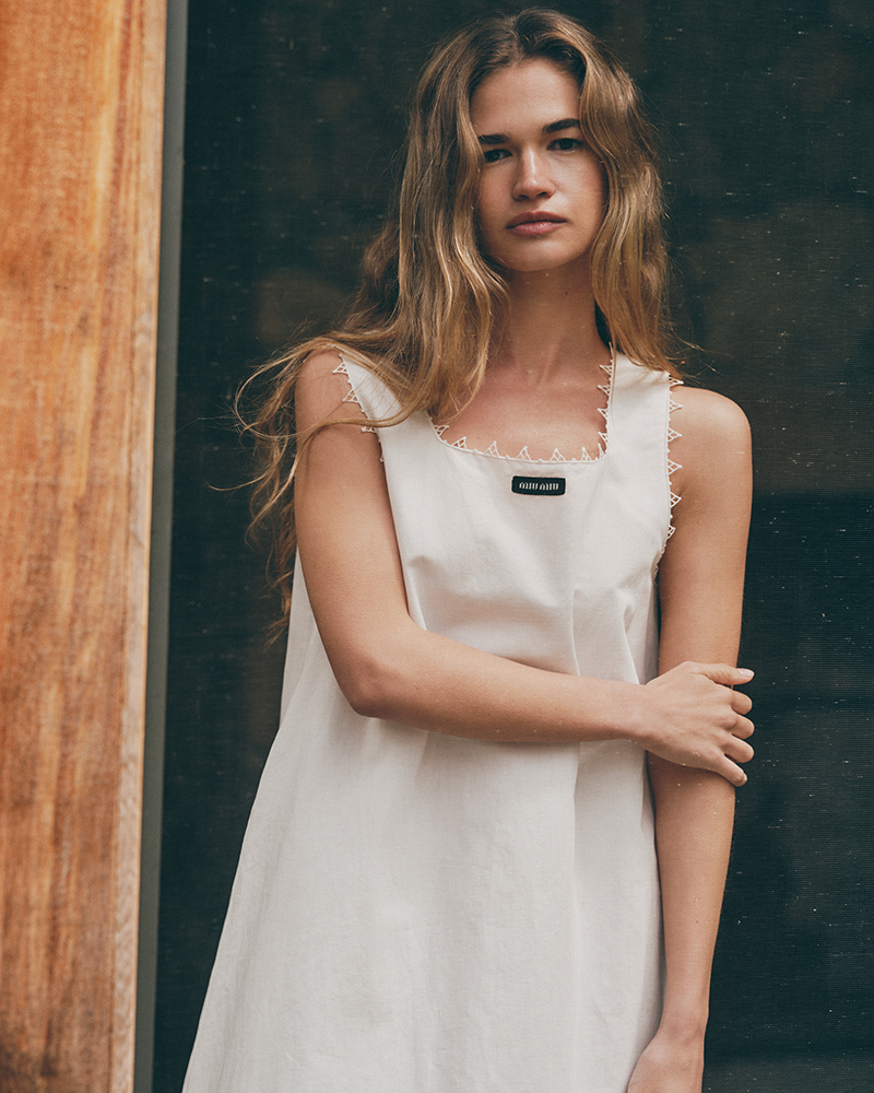 White sleeveless summer dress with delicate trim details%2C styled against a warm-toned outdoor backdrop