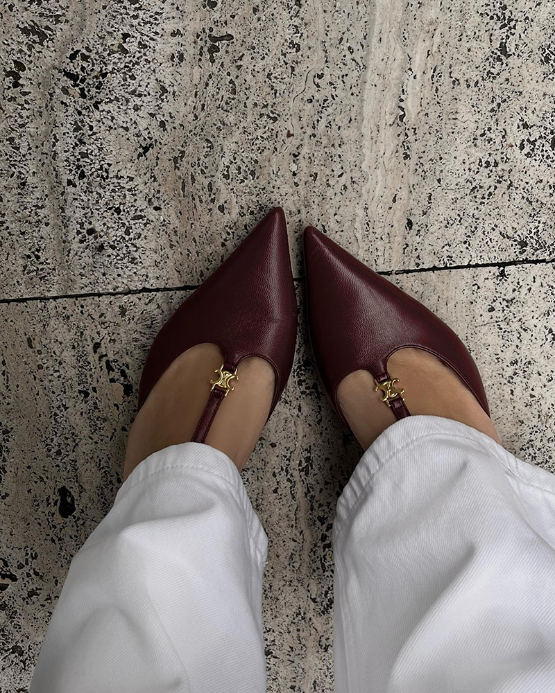 Burgundy leather pointed-toe flats with gold logo hardware styled with white wide-leg trousers on stone flooring