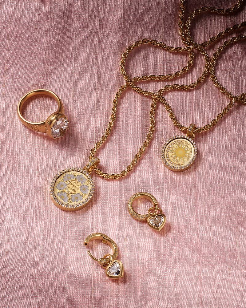 Gold medallion necklaces%2C heart charm earrings%2C and diamond ring styled on soft pink silk backdrop