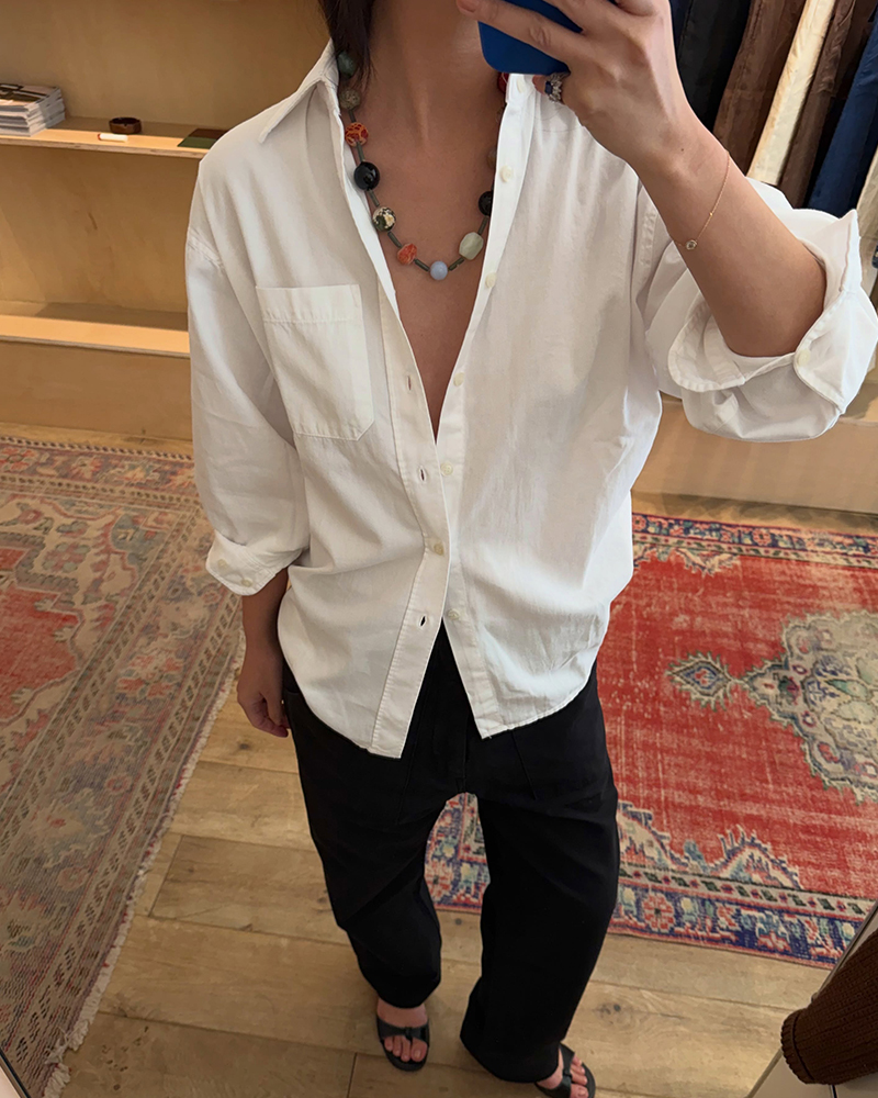 Oversized white button-down shirt styled with black trousers%2C sandals and beaded necklace indoors