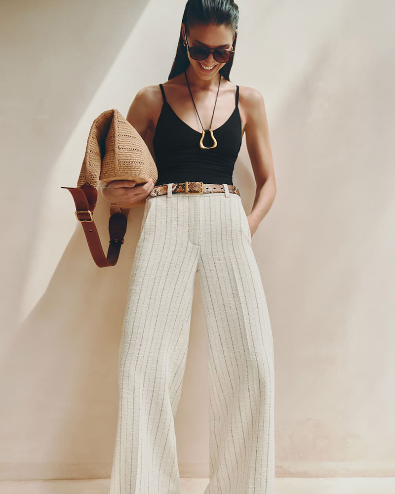 Black camisole bodysuit with striped wide-leg trousers%2C woven shoulder bag and sunglasses in a sunlit resort setting