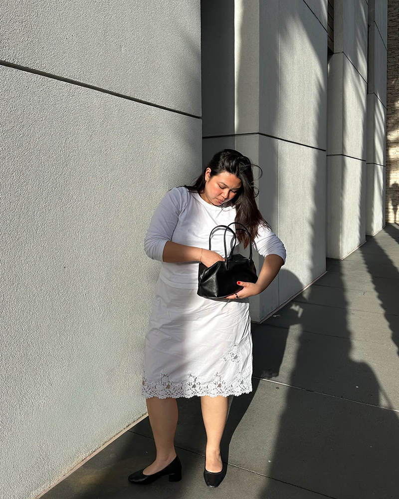 White lace-trim midi dress styled with black leather handbag and pumps against a sunlit city wall
