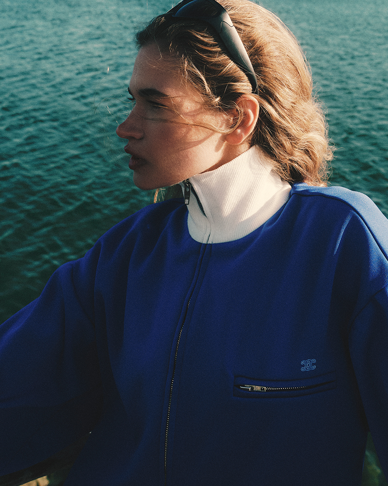 Model in oversized cobalt blue zip-up jacket and white turtleneck sailing on open water in a sporty luxury look