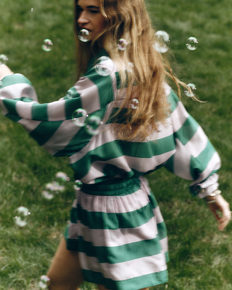 Model wearing a green and white wide striped summer dress with long sleeves and elastic waist%2C surrounded by floating bubbles on grass background
