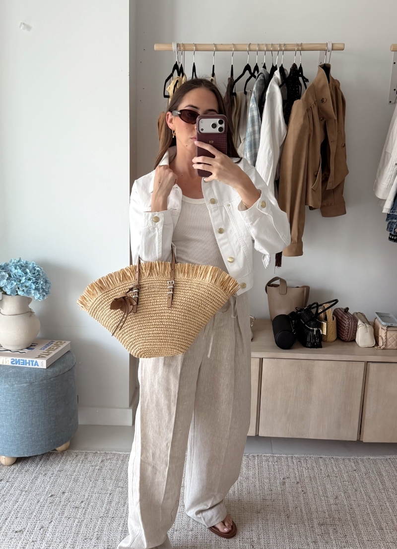 Mirror selfie in a white denim jacket and linen trousers with a woven straw tote in a neutral dressing room