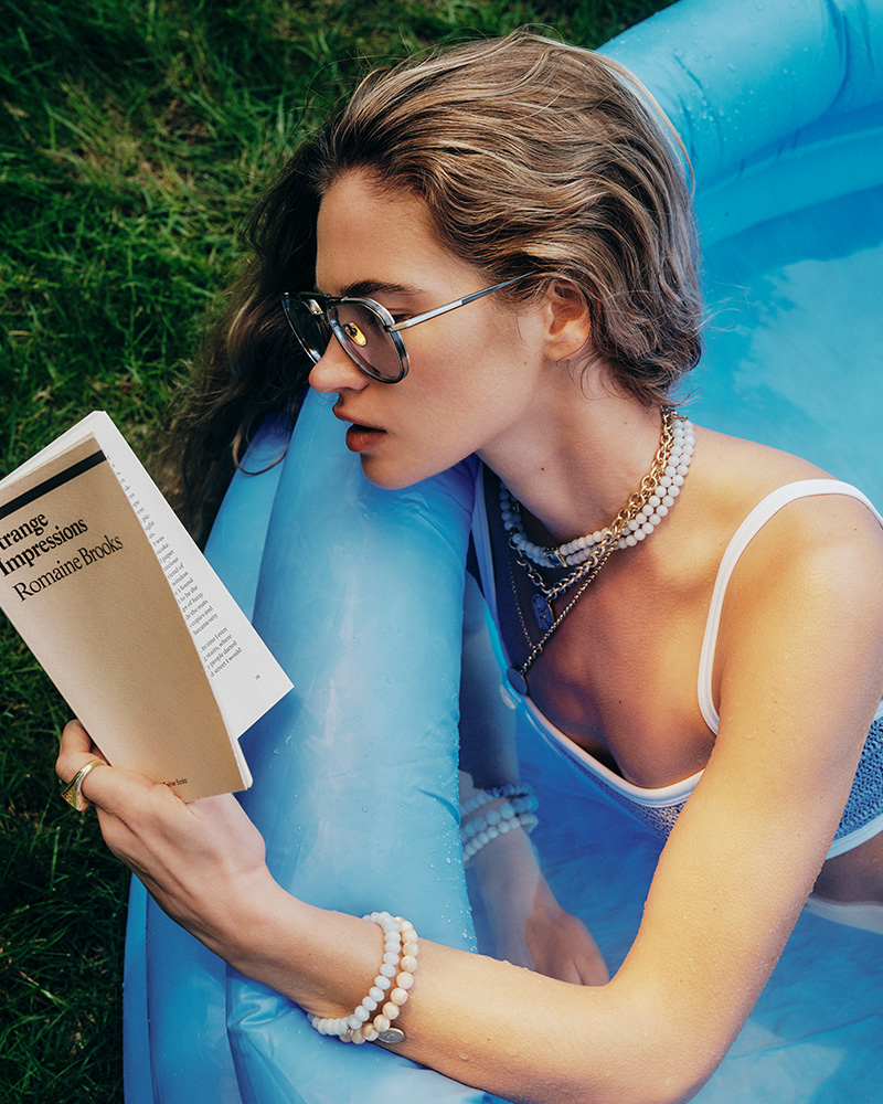 Oversized aviator sunglasses with layered pearl jewelry and blue swimsuit styling beside a poolside inflatable raft