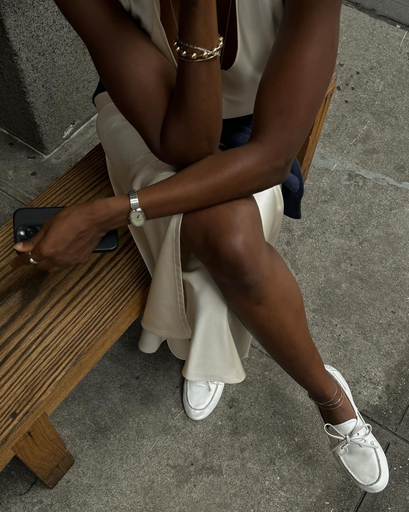 Close-up of cream satin midi dress with white leather loafers and silver jewelry%2C styled on a city bench