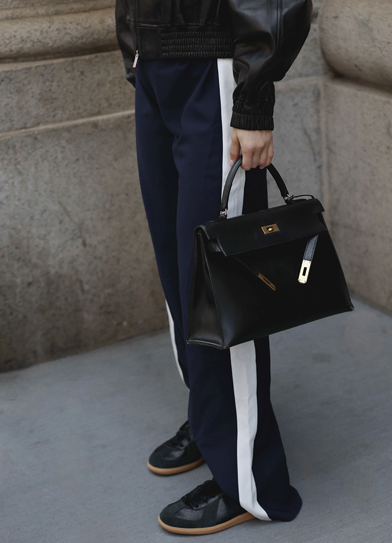 Black leather bomber jacket with striped track pants, oversized tote bag and sneakers on a city street backdrop