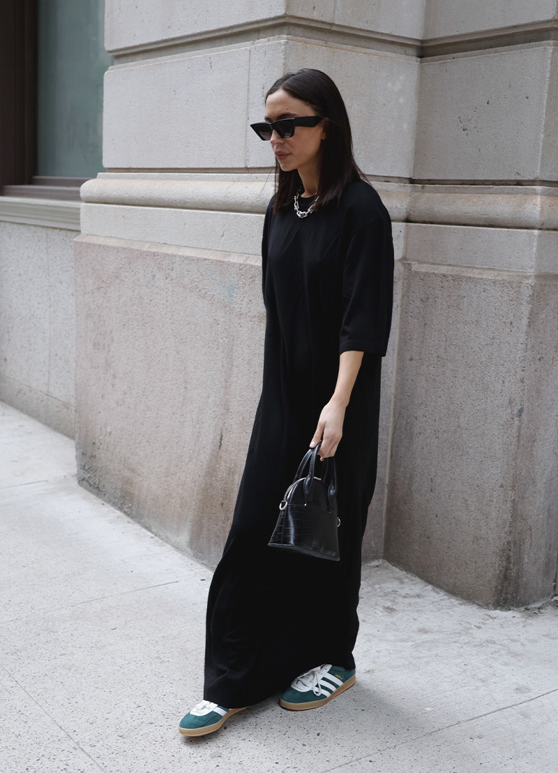 Black maxi dress with angular sunglasses, mini leather handbag and retro sneakers on a city sidewalk backdrop