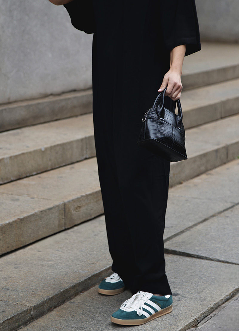 Black maxi dress with angular sunglasses, mini leather handbag and retro sneakers on a city sidewalk backdrop