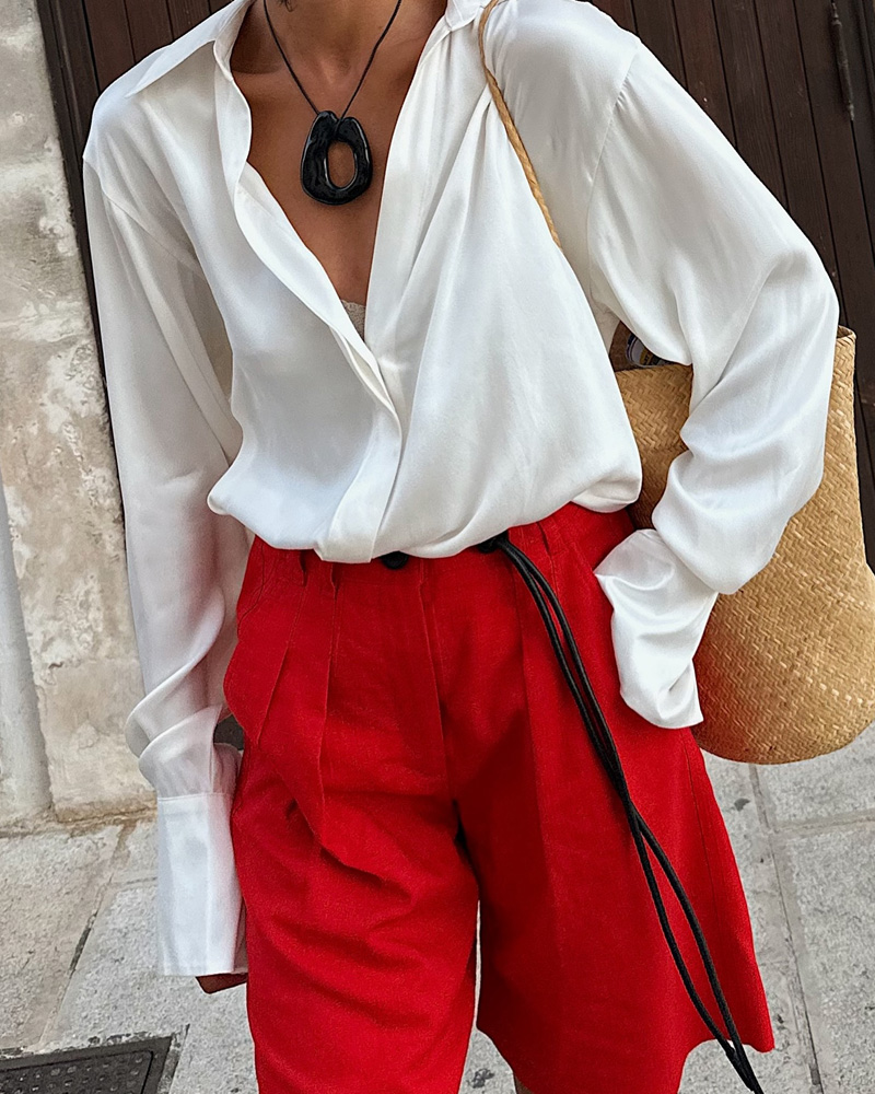 White silk blouse styled with red tailored shorts%2C woven tote bag%2C and flip-flops against a rustic city backdrop