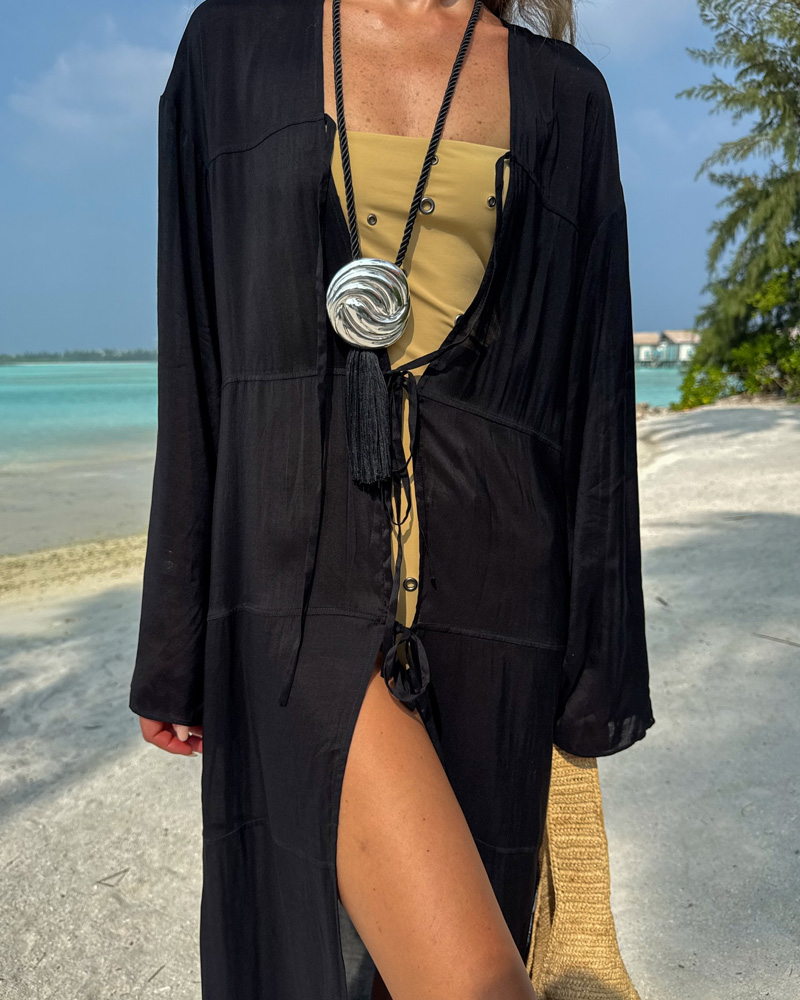 Black sheer beach cover-up layered over a tan swimsuit with a woven tote and statement necklace by the ocean