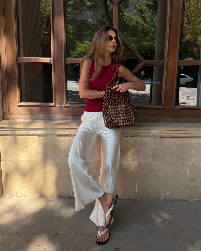 Chic summer outfit featuring a red fitted tank top paired with flowy white satin pants and stylish flip-flops%2C complemented by a brown woven bag.