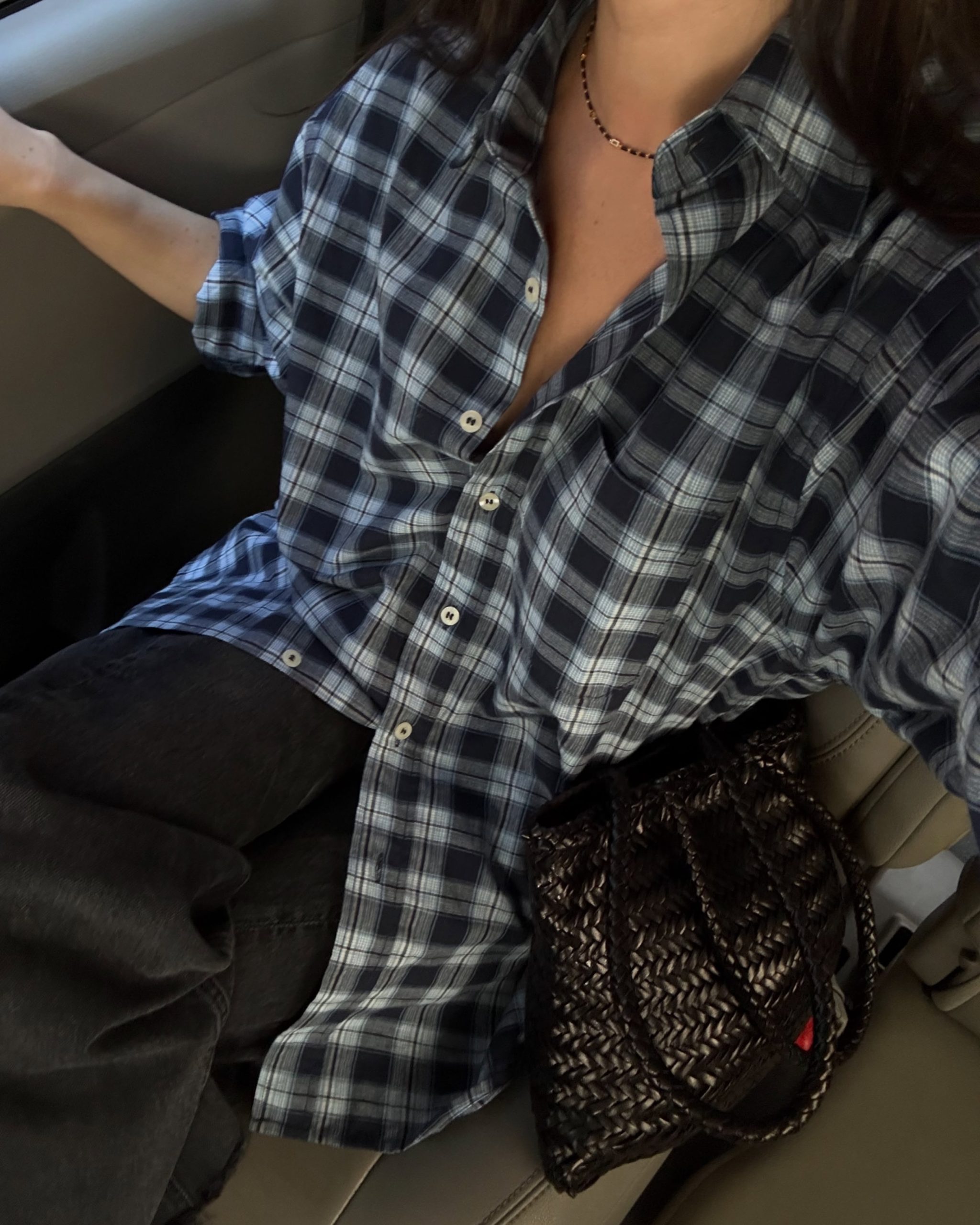 Oversized blue and gray plaid button-up shirt paired with black denim jeans and a woven dark brown leather handbag on a car seat.