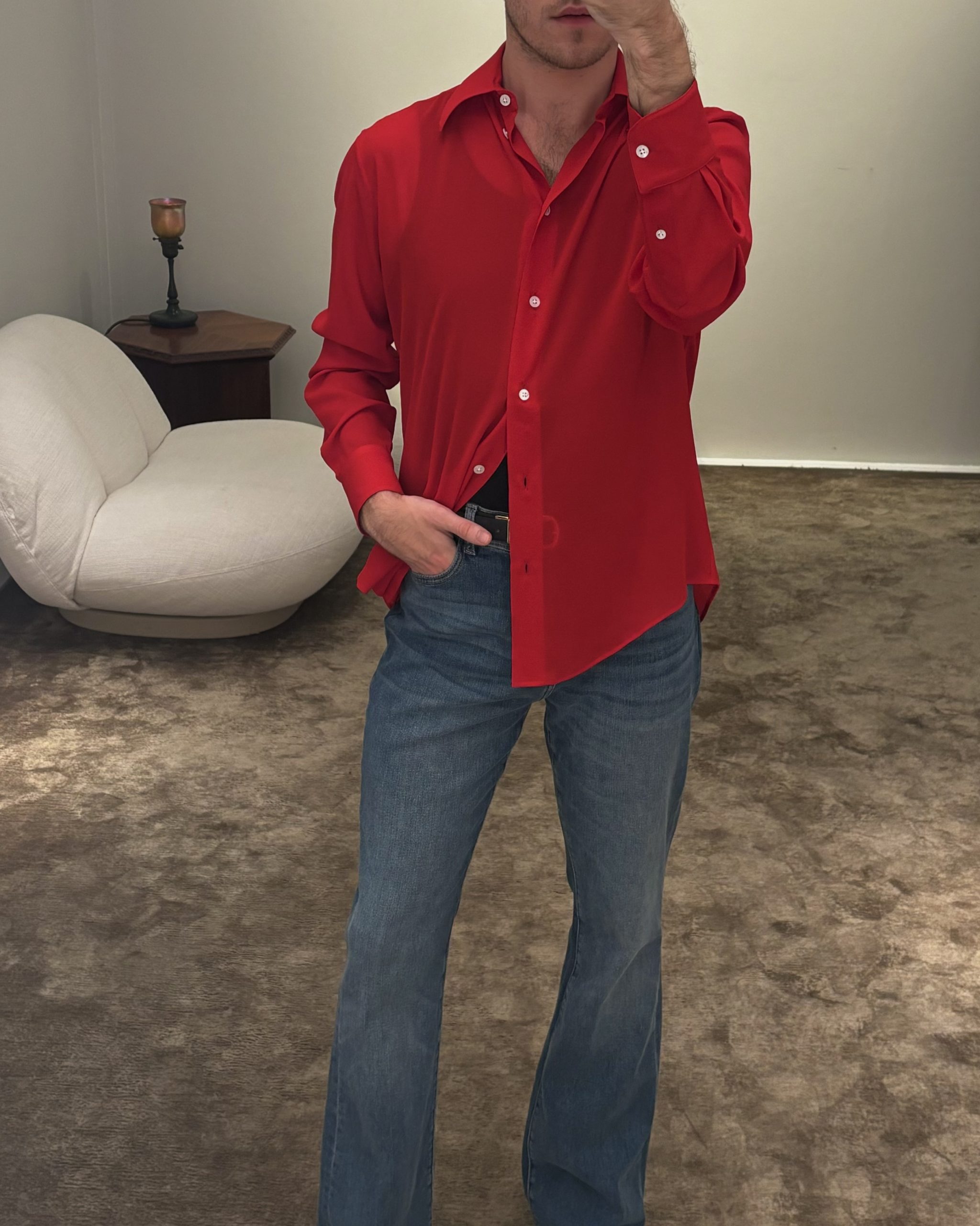 Man wearing a bright red silk button-up shirt paired with high-waisted blue denim jeans in a minimalist room setting.