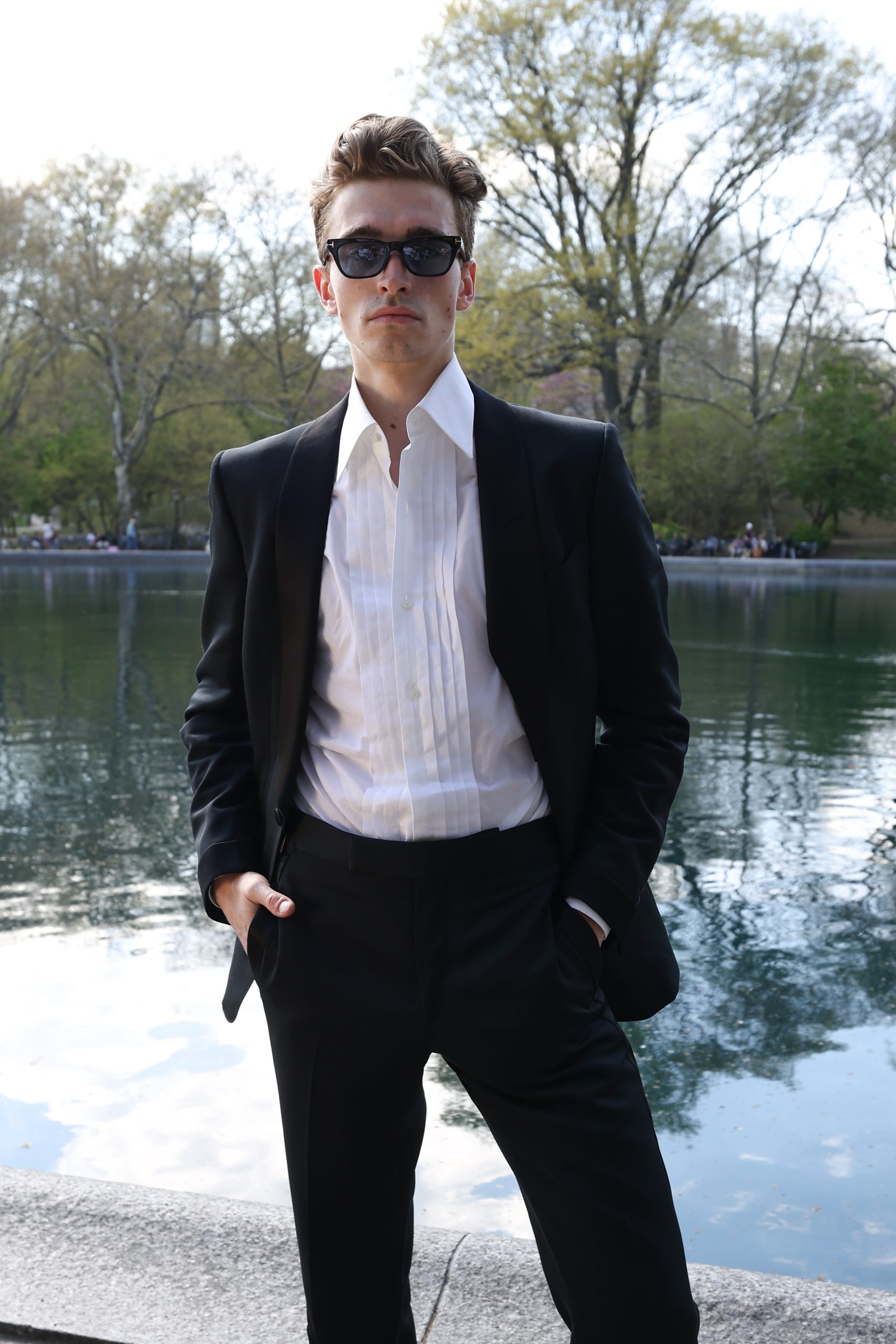 Young man in a black suit and white shirt stands confidently with hands in pockets by a serene pond%2C wearing sunglasses. Trees reflect in the water.