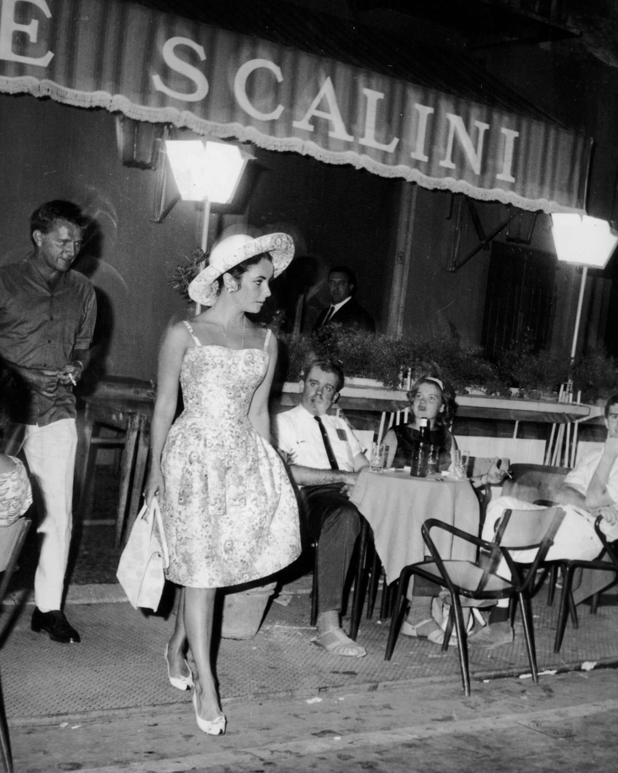 Elegant woman in a floral dress and wide-brimmed hat walks past people seated at an outdoor caf%C3%A9. The ambiance is vintage and glamorous.