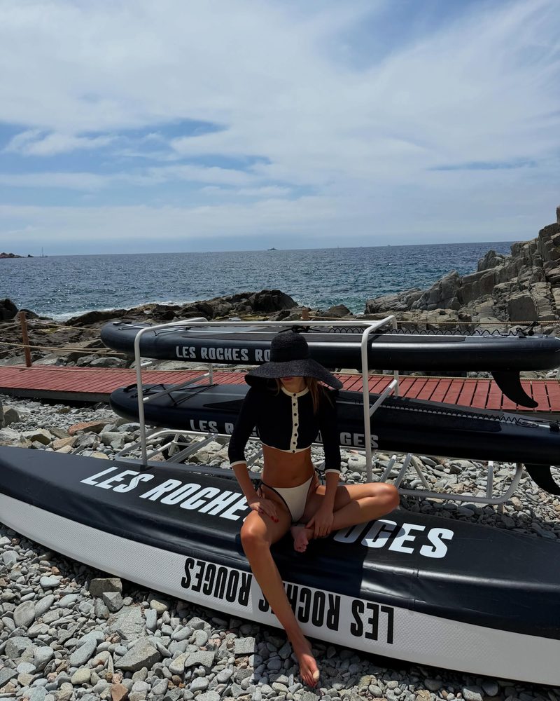 A woman wearing a black sun hat and swimwear sits on a black paddleboard labeled %22Les Roches Rouges%2C%22 against a rocky beach and ocean backdrop.