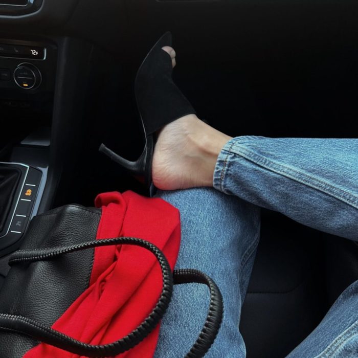 A person wearing jeans and black heeled sandals rests their foot on a car seat. A black handbag and a red garment are nearby, creating a casual and stylish scene.