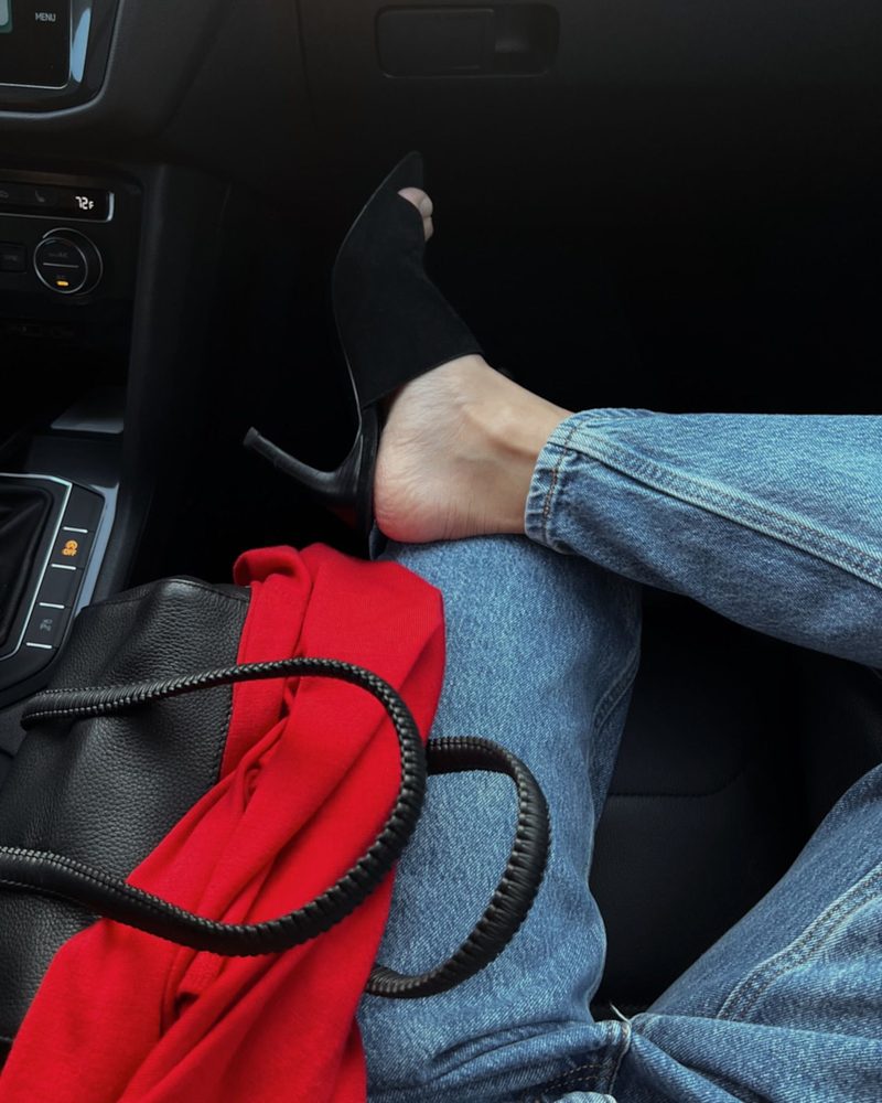 A person wearing jeans and black heeled sandals rests their foot on a car seat. A black handbag and a red garment are nearby%2C creating a casual and stylish scene.