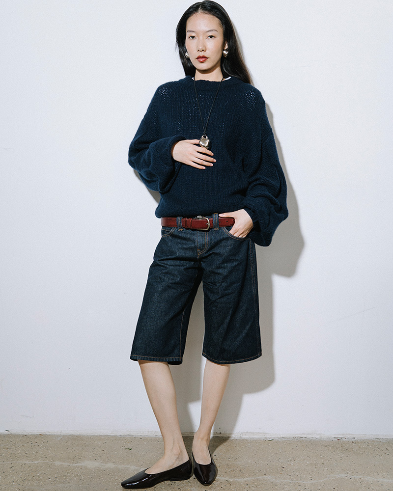 Navy oversized sweater paired with dark denim Bermuda shorts%2C a brown belt and pendant necklace in studio