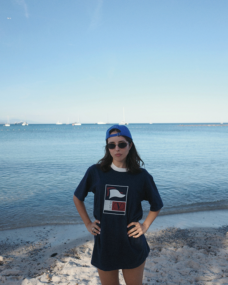 Vintage graphic T-shirt with a blue baseball cap and slim sunglasses%2C styled on a sandy beach by calm ocean waters