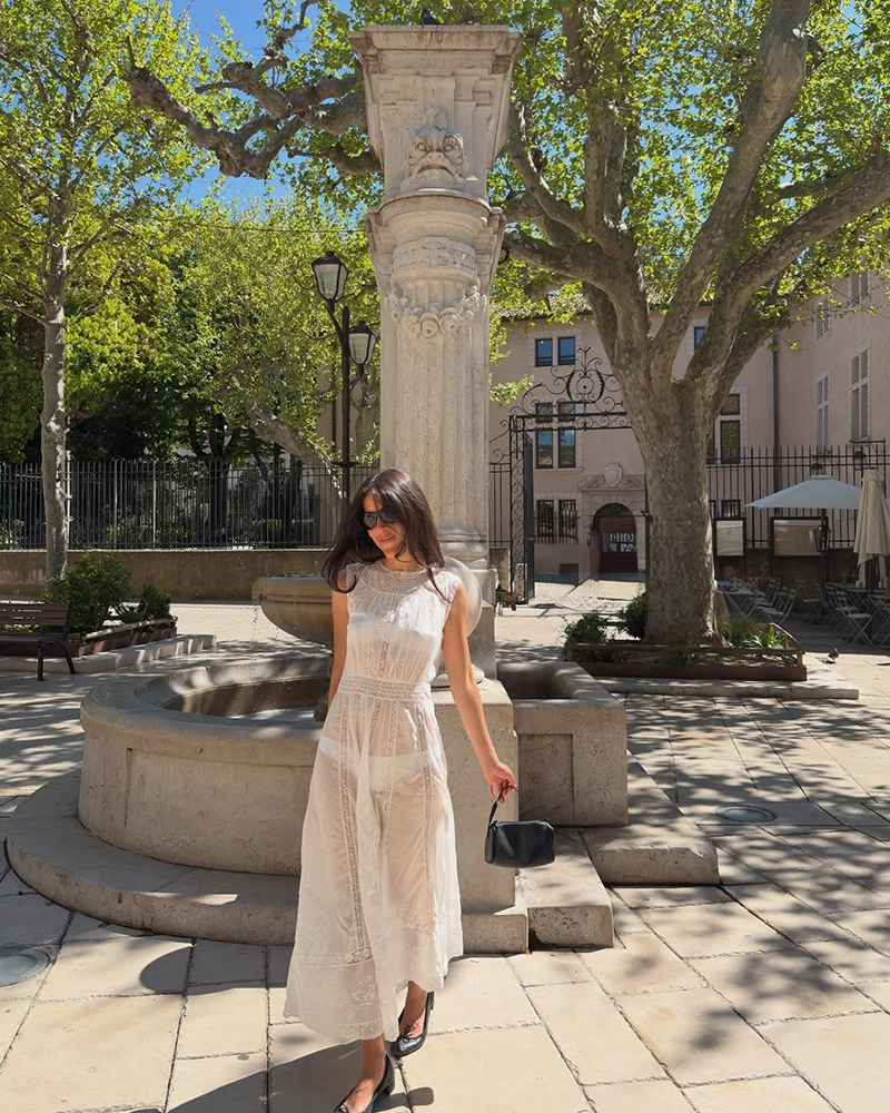 White lace midi dress with black heeled ballerinas%2C sunglasses%2C and a mini black handbag in a sunlit European square by a stone fountain