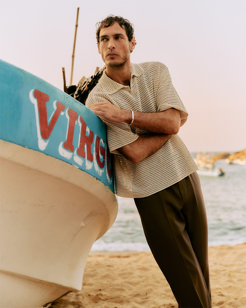 Men%E2%80%99s summer outfit featuring a beige open-knit polo shirt and tailored trousers%2C styled beside a fishing boat on a sandy beach backdrop