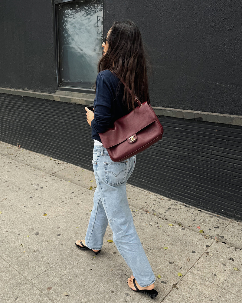 Chanel burgundy leather flap shoulder bag paired with a navy sweater%2C light-wash jeans and sandals on a city sidewalk backdrop