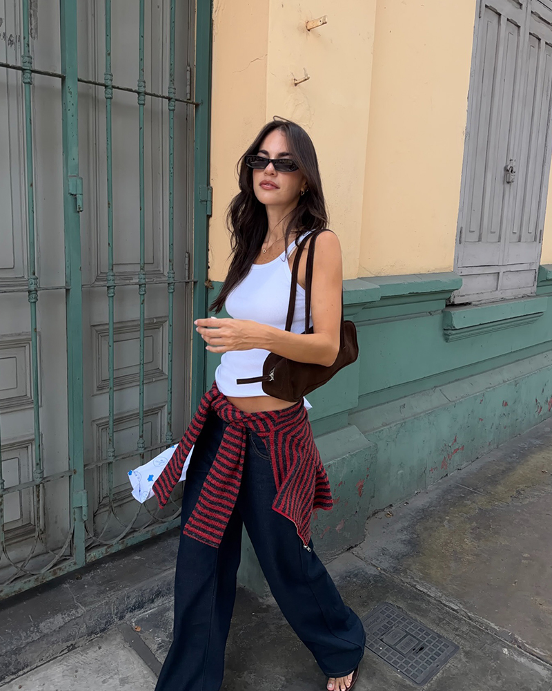 Brunette in a white ribbed tank top%2C wide-leg navy trousers and brown shoulder bag on a pastel city street backdrop