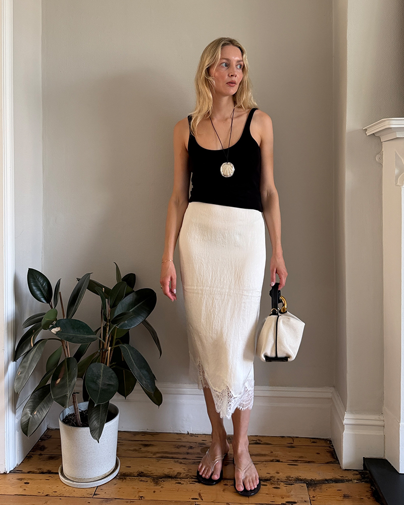 Black tank top paired with a cream lace-trim midi skirt%2C mini top-handle bag and flip-flops in a minimalist interior setting