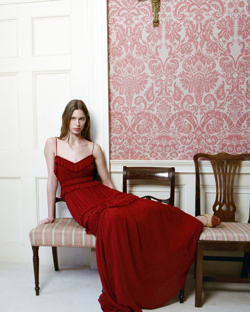 Model in a red ruffled maxi dress and strappy heels%2C seated against ornate pink wallpaper in an elegant interior
