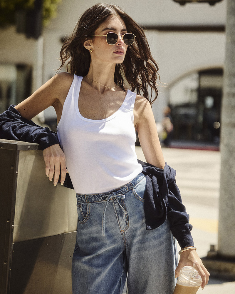 Woman in a white tank top and relaxed blue jeans with sunglasses and a navy cardigan%2C styled on a sunlit city street
