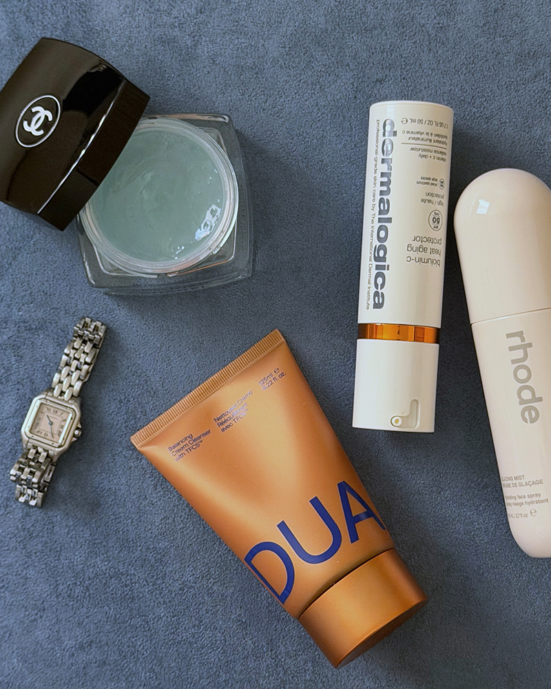 Chanel cream jar%2C Dermalogica sunscreen%2C Rhode mist and DUA body cream with silver watch on blue fabric backdrop