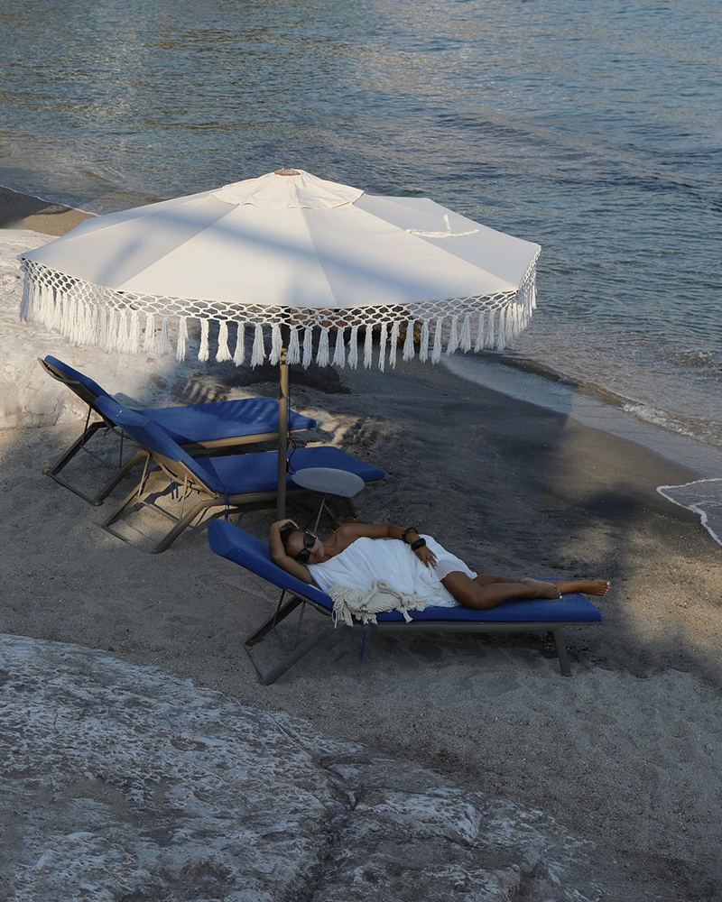 White beach cover-up and sunglasses styled on a seaside sun lounger beneath a fringed umbrella on a Mediterranean shore
