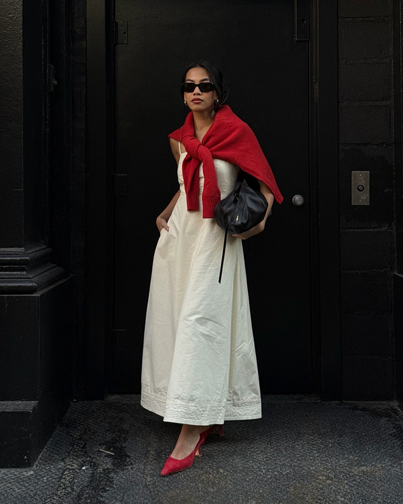 White sleeveless maxi dress styled with a red sweater%2C black shoulder bag and red pumps against a dark city doorway