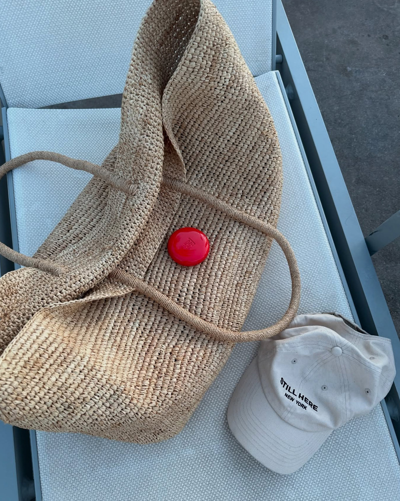 Oversized woven raffia tote with a white Still Here New York cap%2C styled on a poolside lounge chair for summer travel