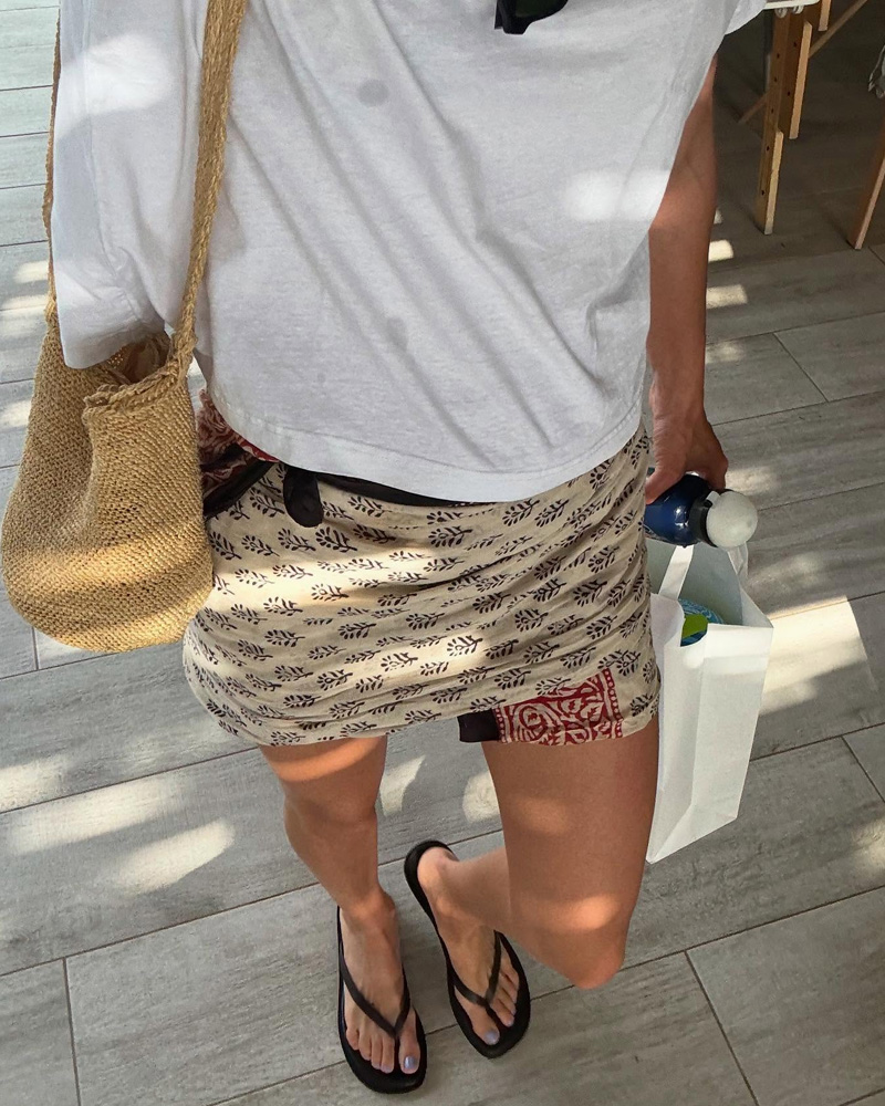 Casual summer outfit featuring a white tee%2C printed mini skirt and black thong sandals%2C styled with a woven shoulder bag indoors