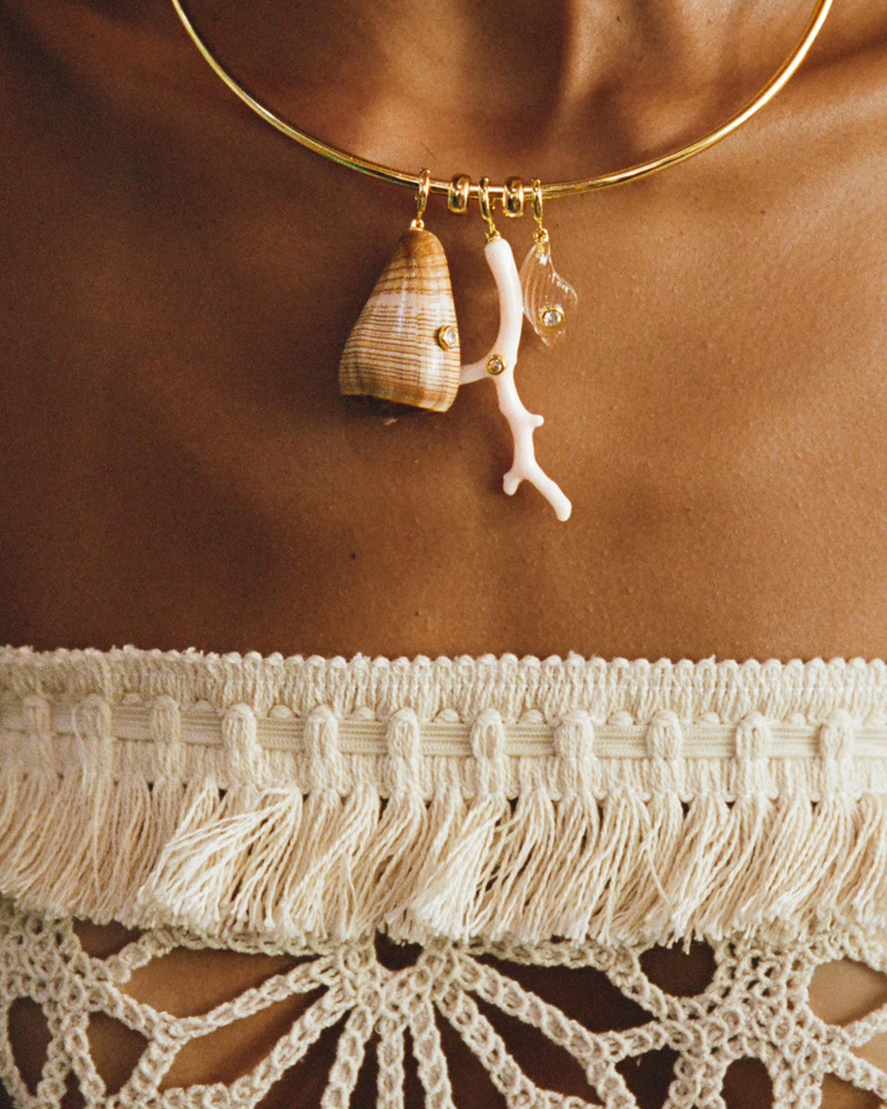 Close-up of a gold charm necklace with shell and coral pendants%2C paired with a cream crochet fringe top in a summer setting