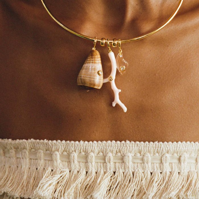 Close-up of a gold charm necklace with shell and coral pendants, paired with a cream crochet fringe top in a summer setting