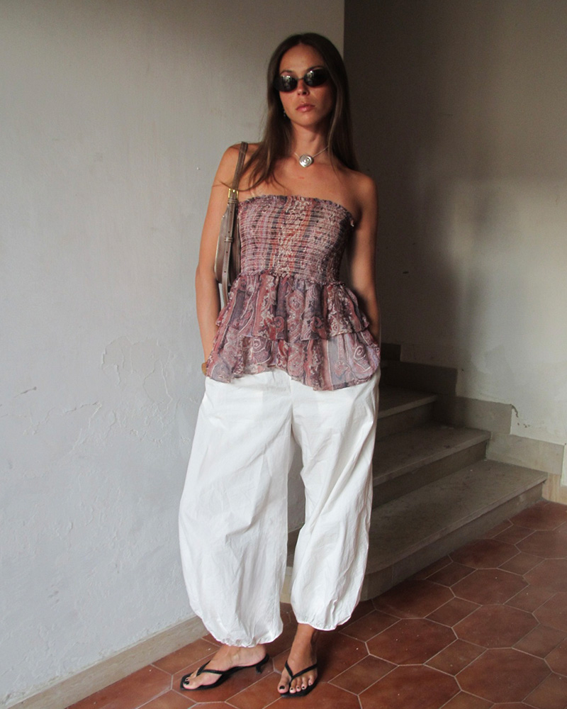 European summer outfit featuring a printed strapless peplum top%2C white balloon trousers%2C sunglasses and shoulder bag in a rustic interior setting