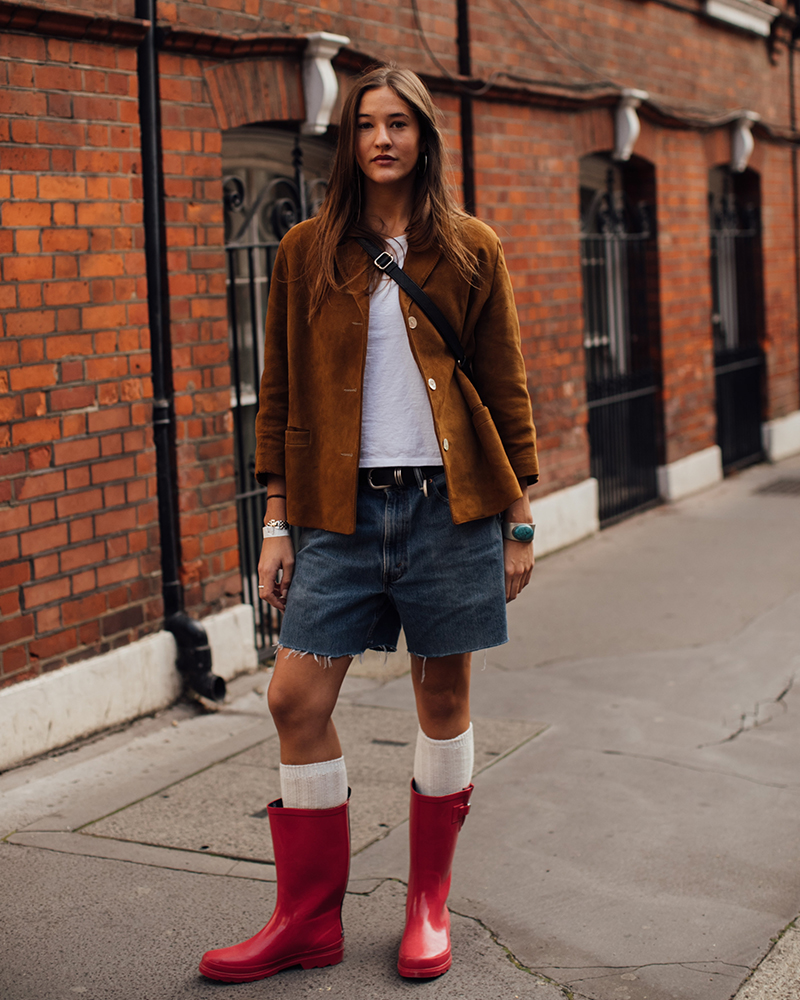 Model in a brown suede blazer%2C denim shorts%2C white socks and red rain boots on a city sidewalk