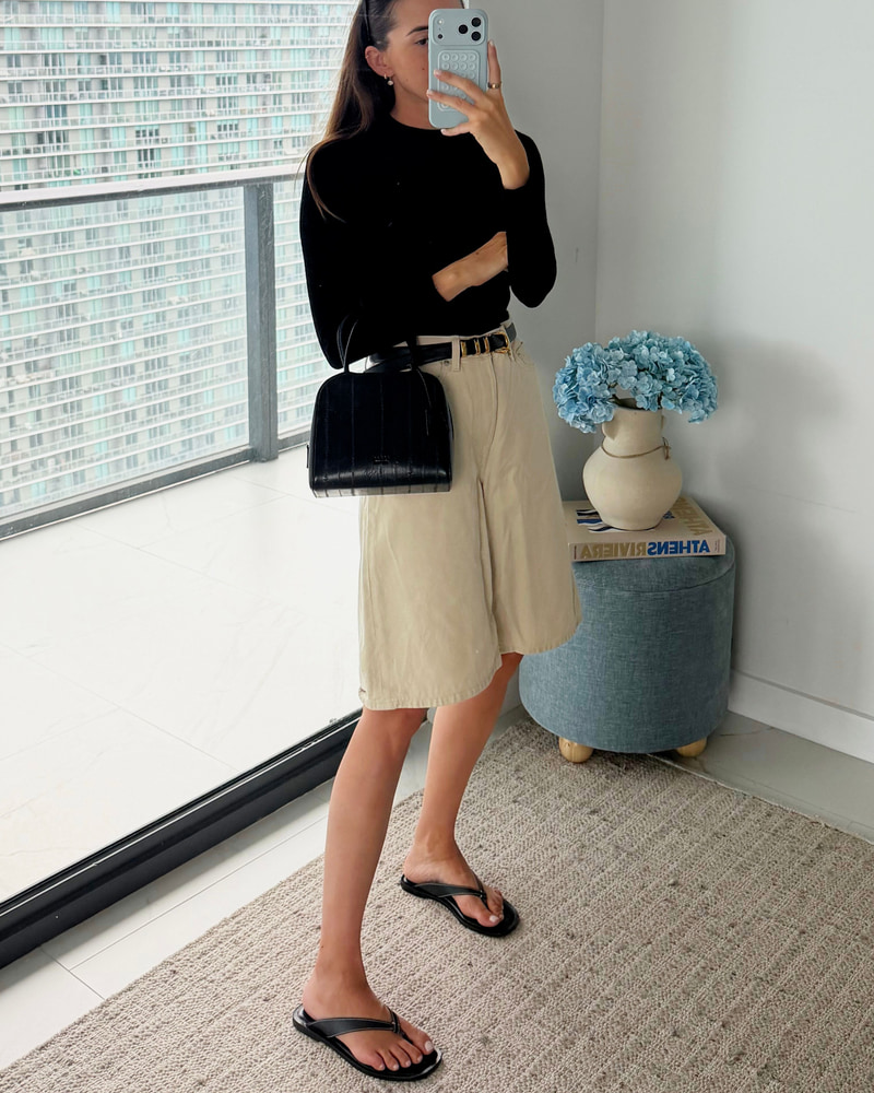 Woman wearing black leather flip-flops paired with beige knee-length shorts%2C black long-sleeve top%2C and a black handbag in a modern apartment setting